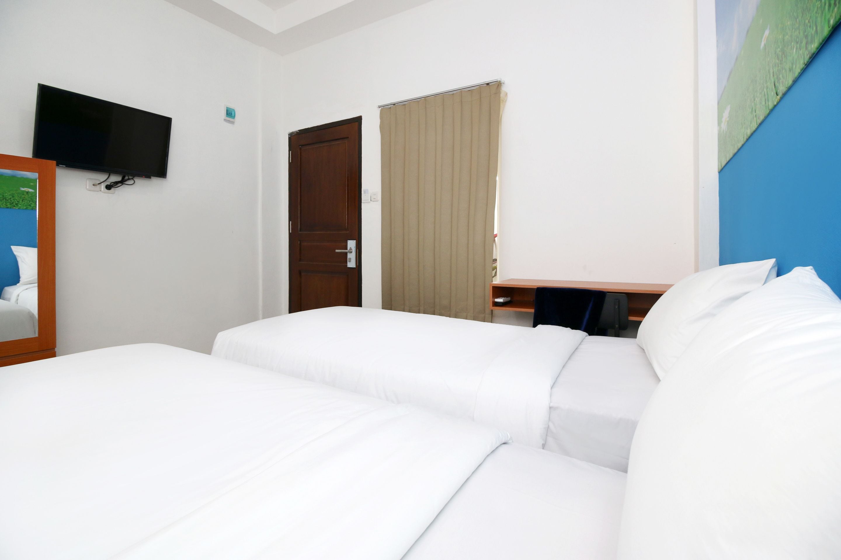 Quarto de hotel com cama, televisão fixada na parede, porta e janela com cortinas.