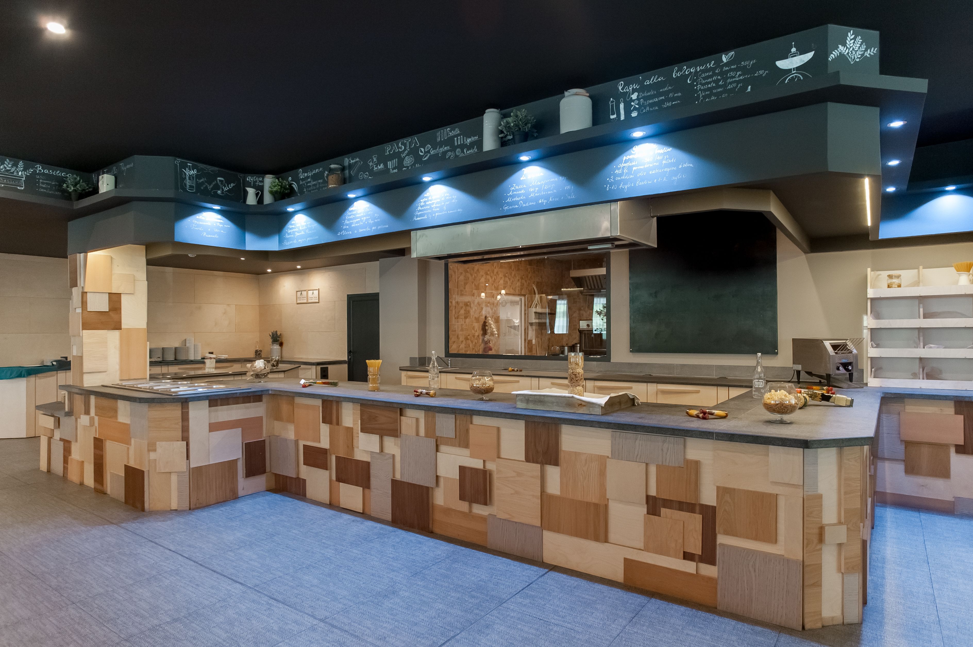 Cozinha moderna com ilha central, detalhes em madeira e um quadro negro exibindo diversos pratos.