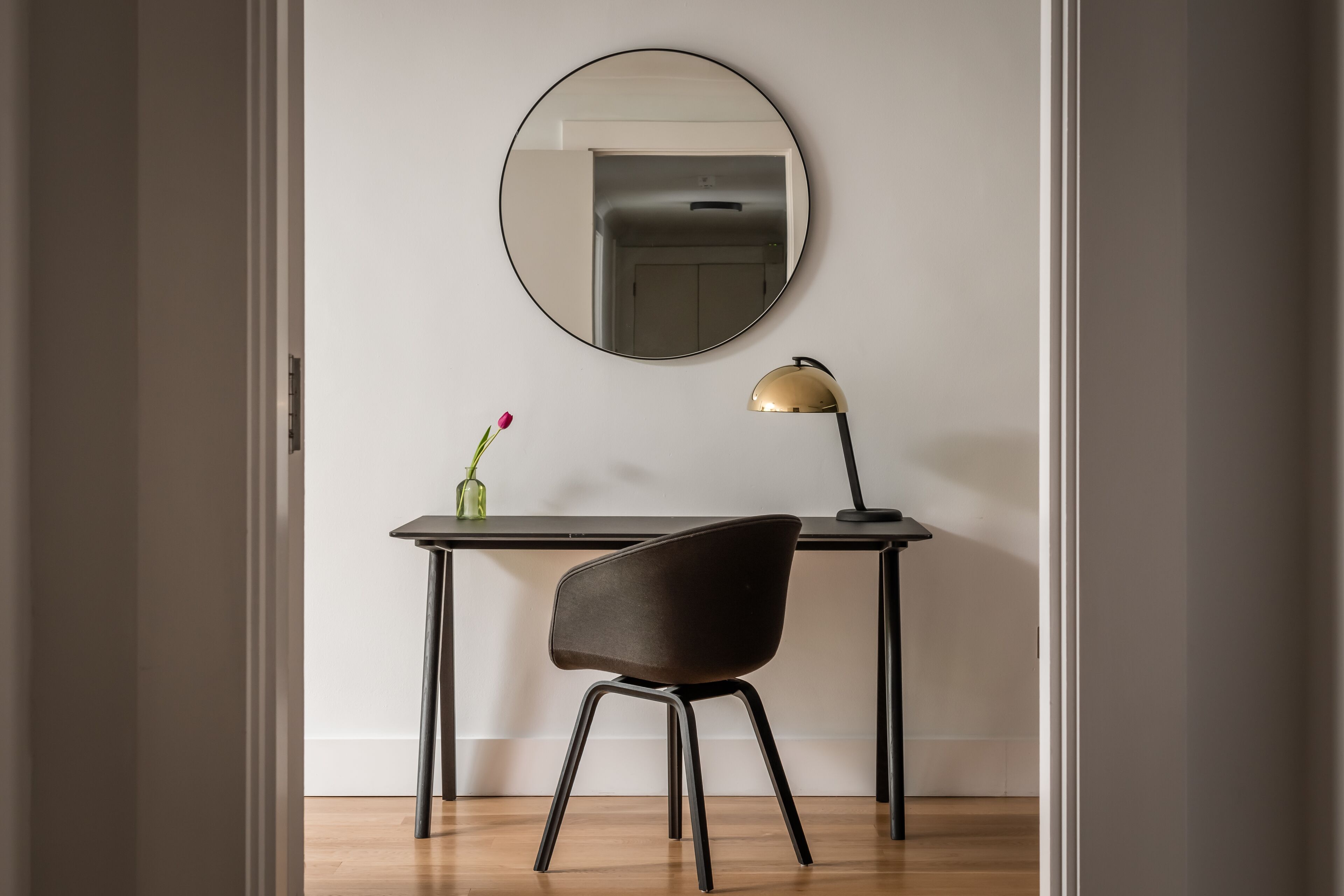 Uma mesa com uma cadeira, um espelho redondo e uma luminária.