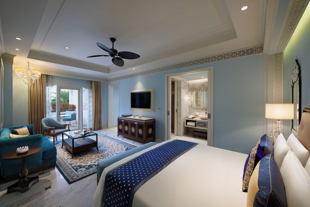 Quarto de hotel com uma cama grande, um sofá, uma mesa de centro, uma TV e um banheiro visível por uma porta aberta.