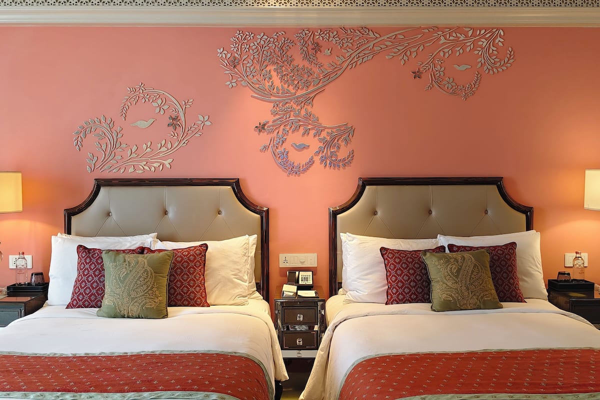 Uma cama de casal com colcha vermelha e branca estampada, duas mesas de cabeceira com luminárias e um detalhe decorativo na parede.