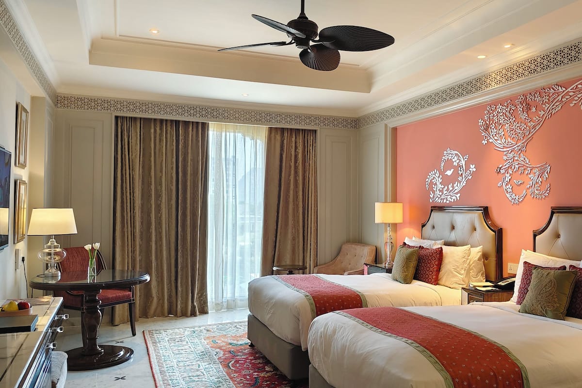 Quarto de hotel com duas camas, ventoinha no teto, parede decorativa e vista pela janela.