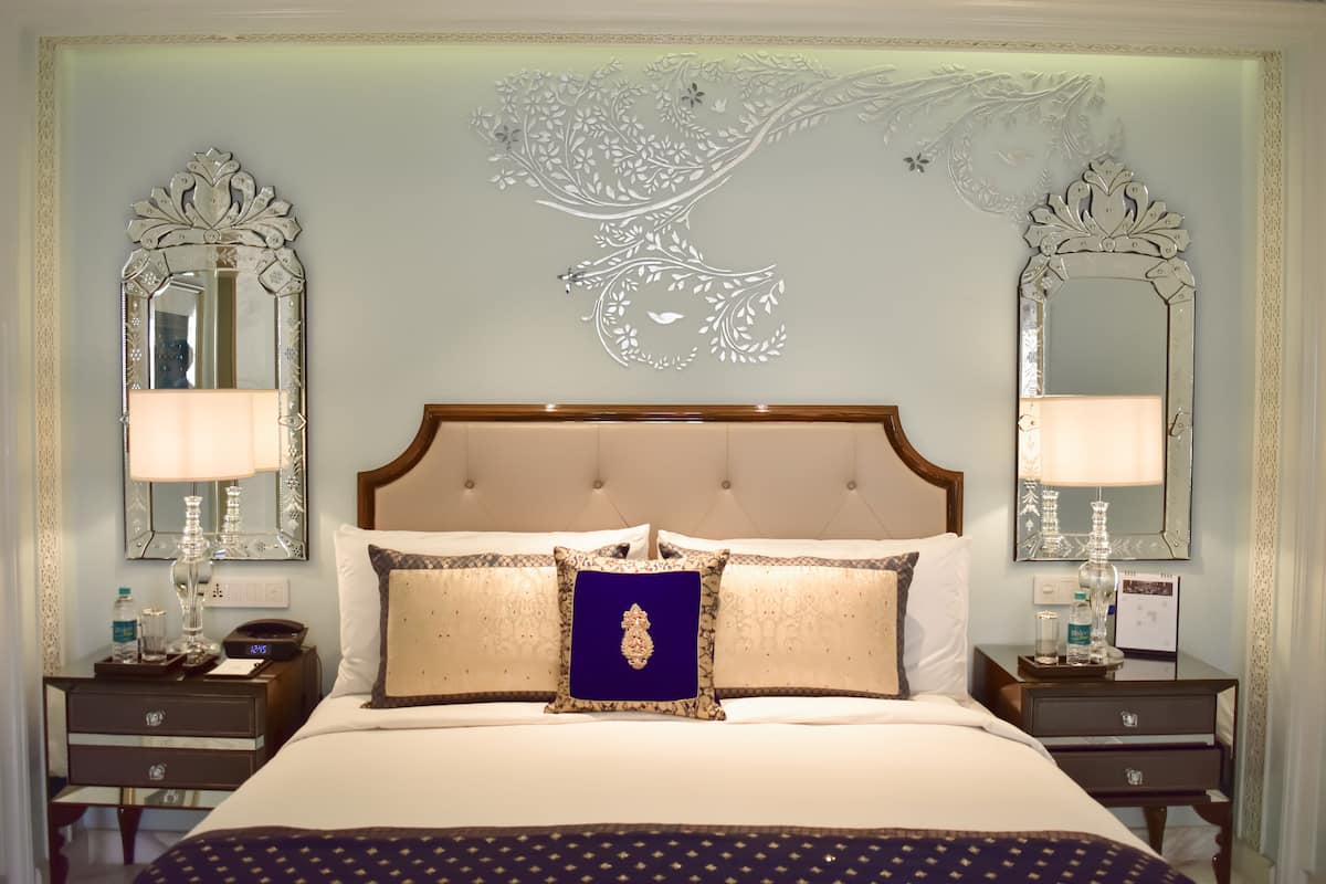 Quarto de hotel com uma cama grande, duas mesas de cabeceira com luminárias, um espelho fixado na parede e um desenho decorativo de árvore na parede.