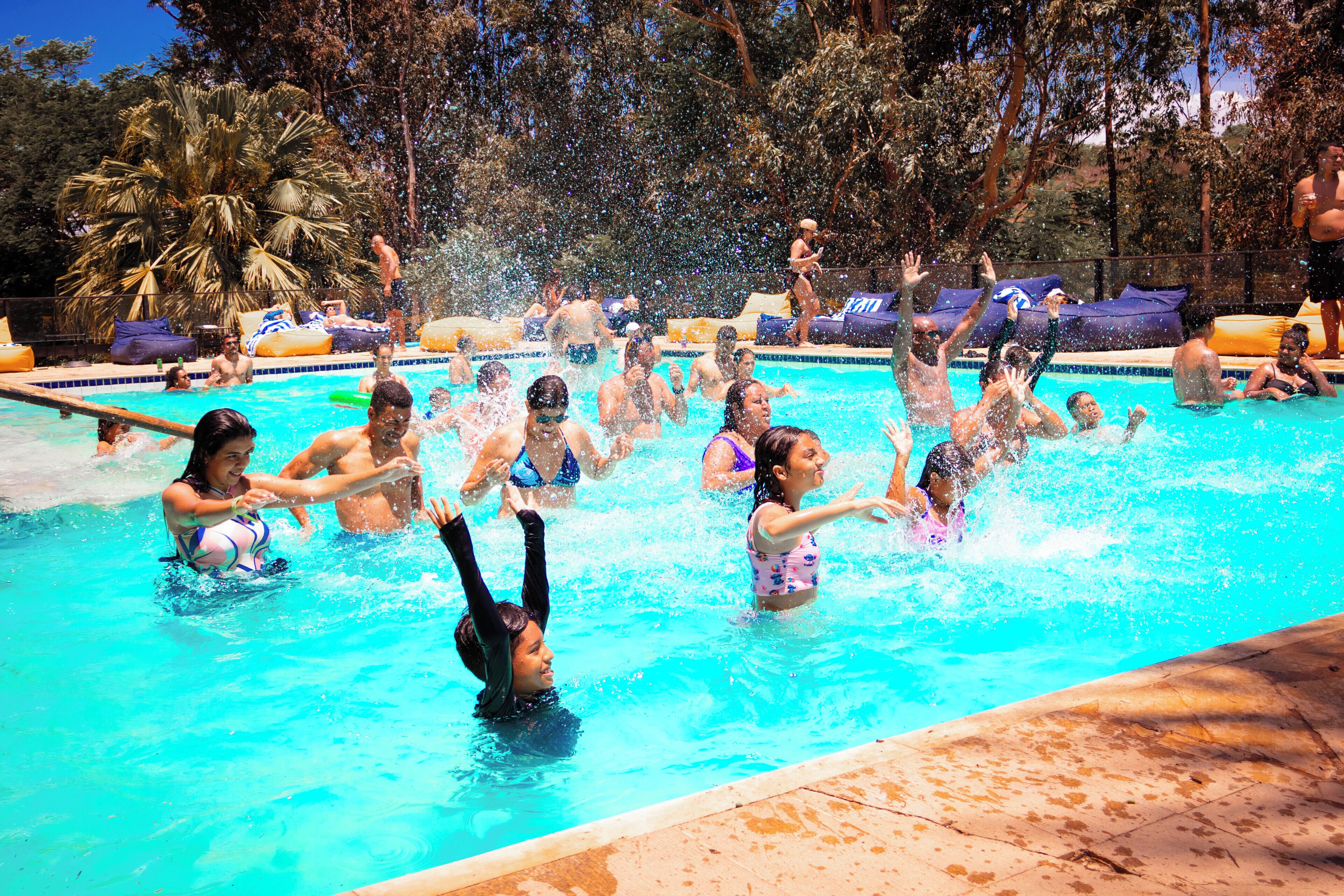 Piscina com pessoas aproveitando a água, cercada por palmeiras e cadeiras de sol.