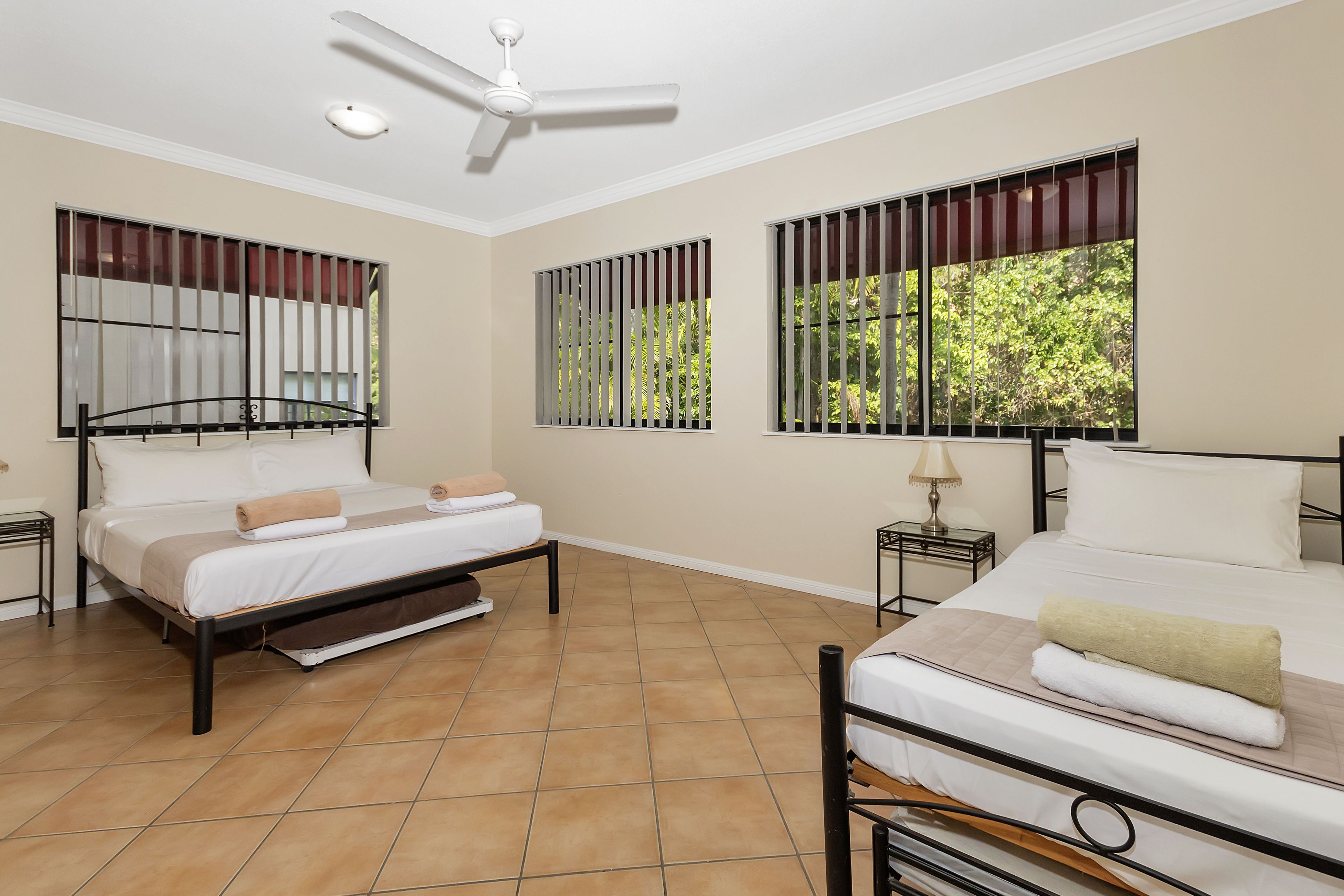 Quarto de hotel com duas camas, ventilador no teto e janelas com grades.