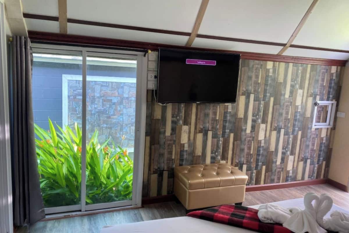 Quarto de hotel com porta de vidro deslizante para uma varanda, TV de tela plana na parede, um banquinho de couro trabalhado e uma cama com um cobertor xadrez vermelho e preto.