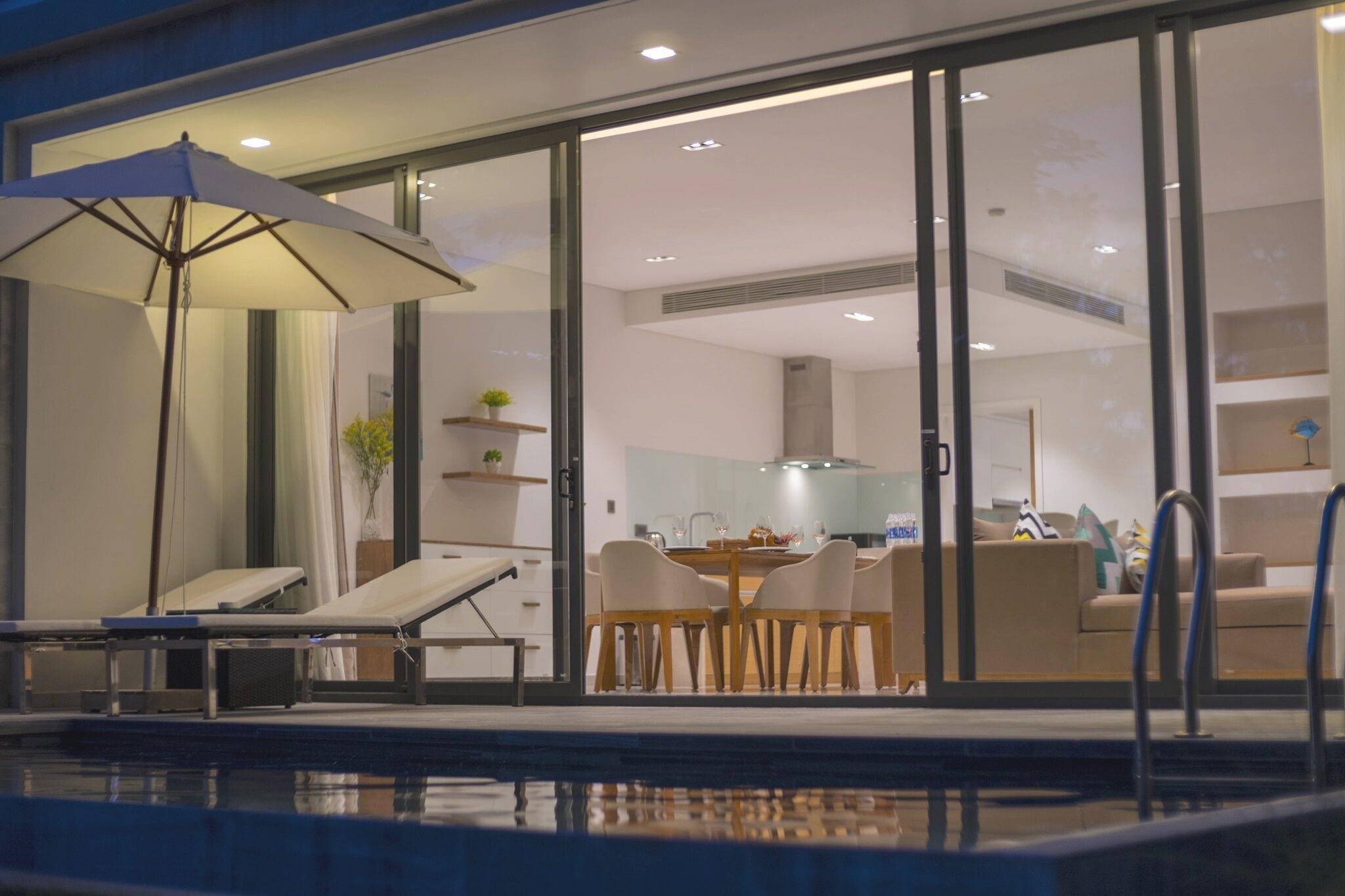 Uma área moderna de piscina interna com espreguiçadeira e guarda-sol, mesa de jantar com cadeiras e cozinha visível através de portas de vidro.