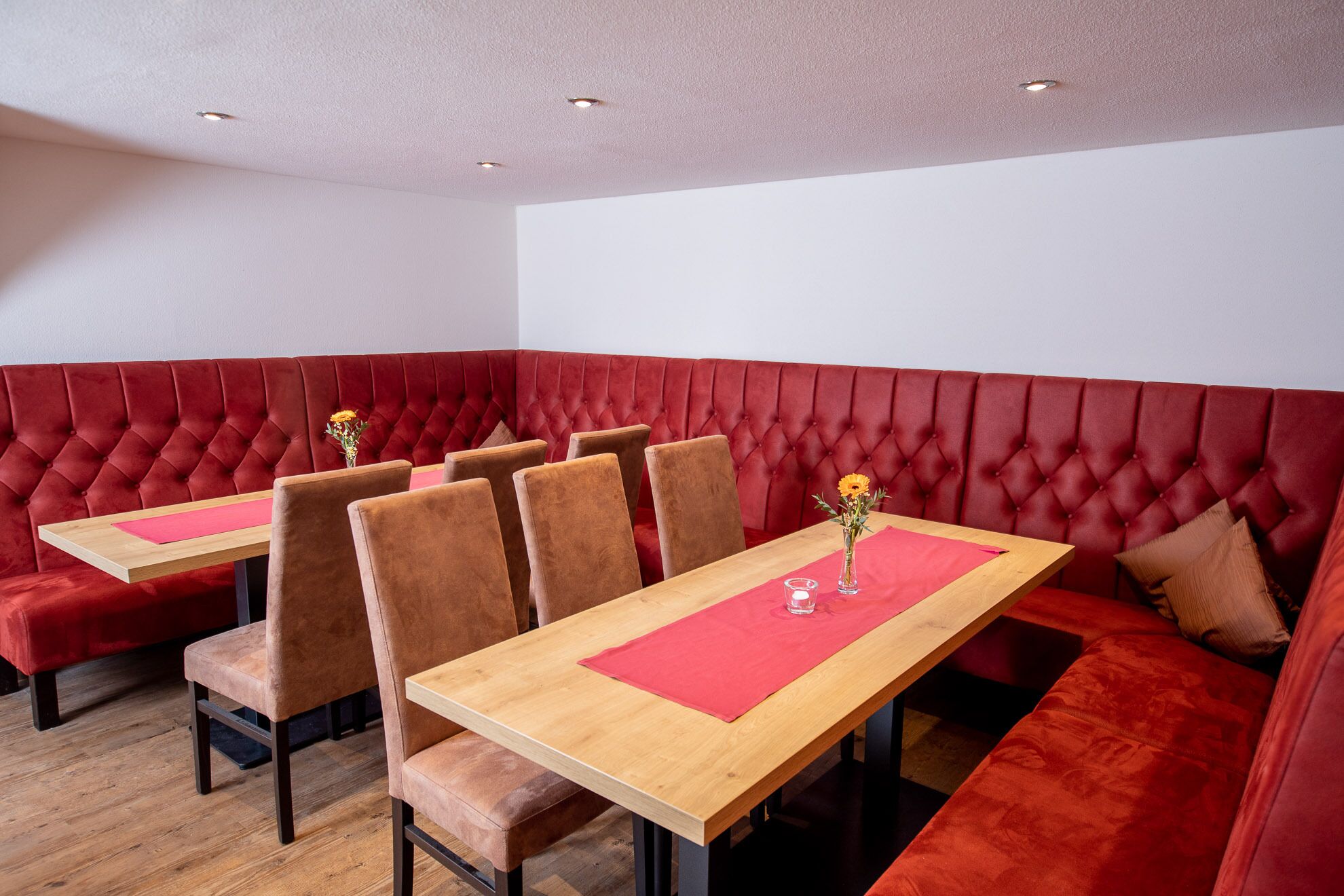 Uma sala de jantar com assentos acolchoados vermelhos e mesas de madeira, cada uma com um jogo americano vermelho e um vaso de flores.