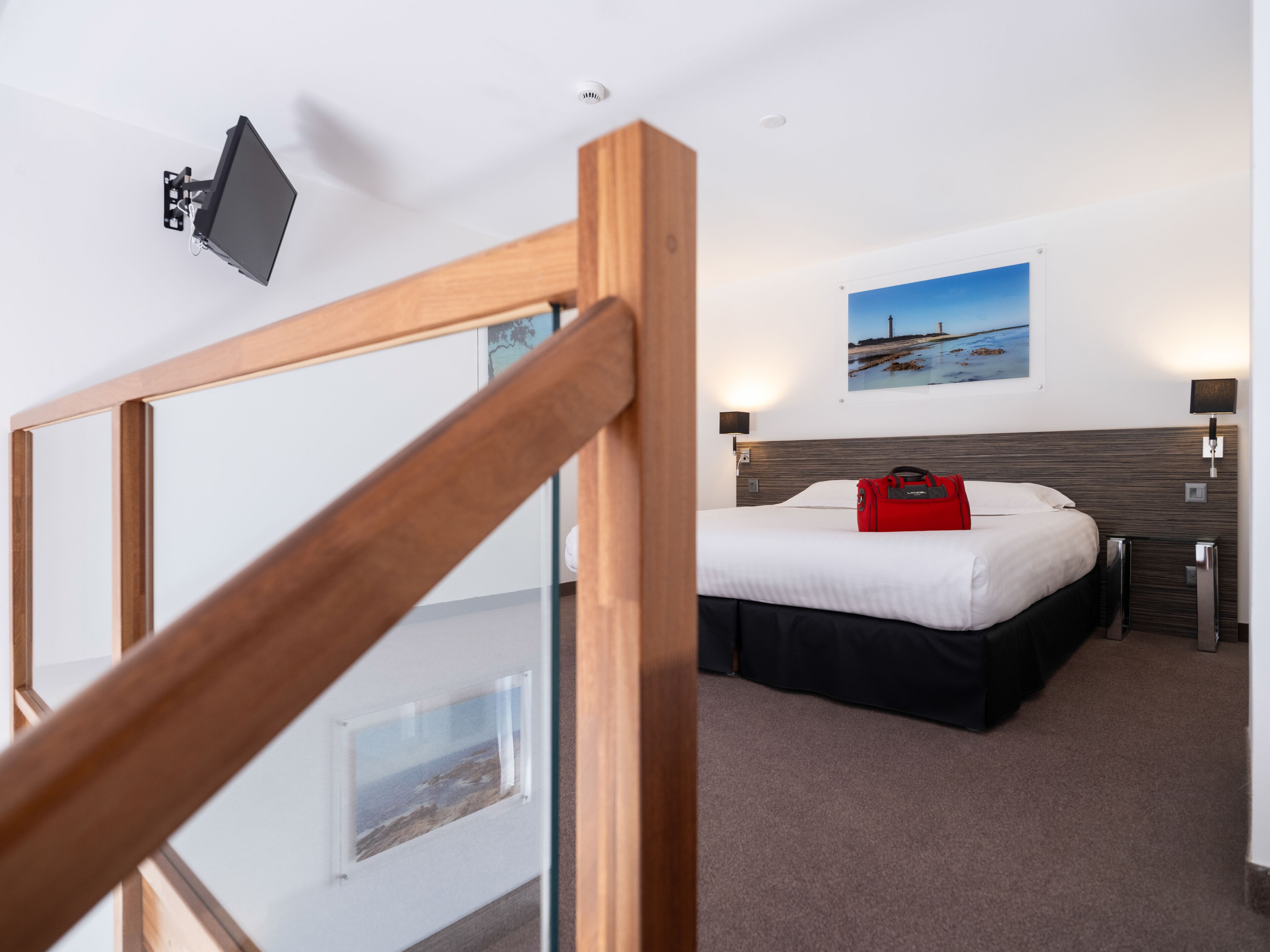Quarto de hotel com cama, abajures, uma mala vermelha e um quadro na parede.