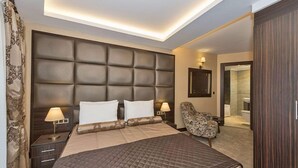 Quarto de hotel moderno com uma cama grande, cabeceira, duas mesas de cabeceira com abajures, uma cadeira e um banheiro visível através de uma porta aberta.