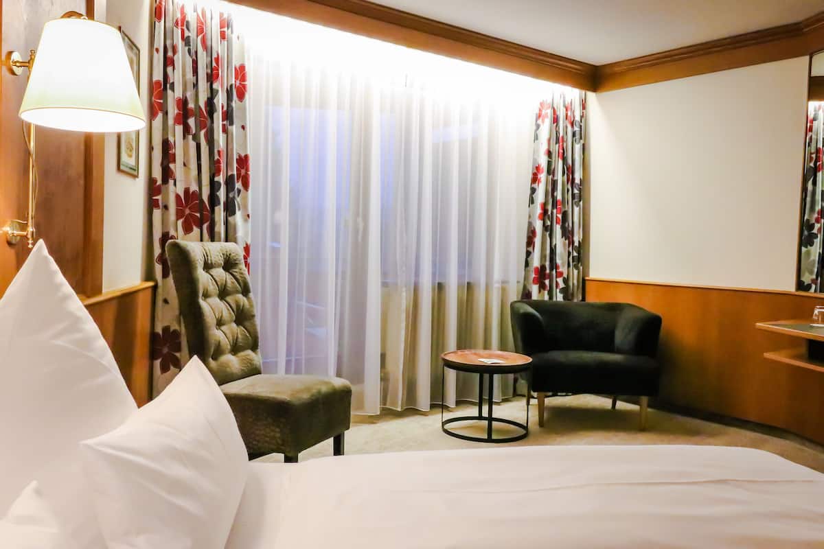 Quarto de hotel com cama, uma cadeira, uma mesinha, abajur e cortinas com padrão floral.