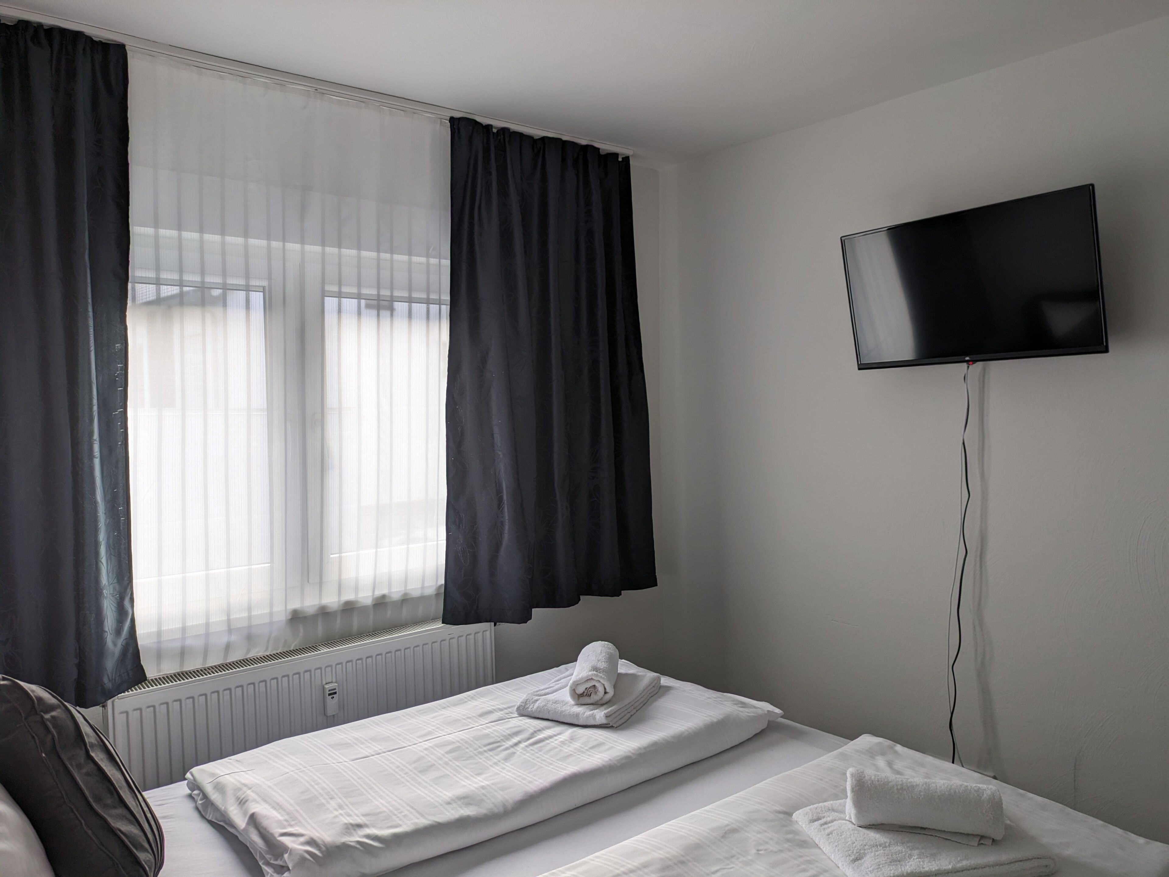 Quarto de hotel com cama, televisão fixada na parede e janela com cortinas.
