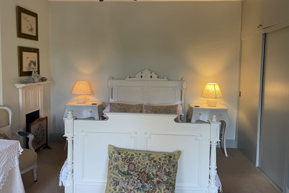 Quarto com cama branca, duas mesas de cabeceira com luminárias, lareira e quadros na parede.