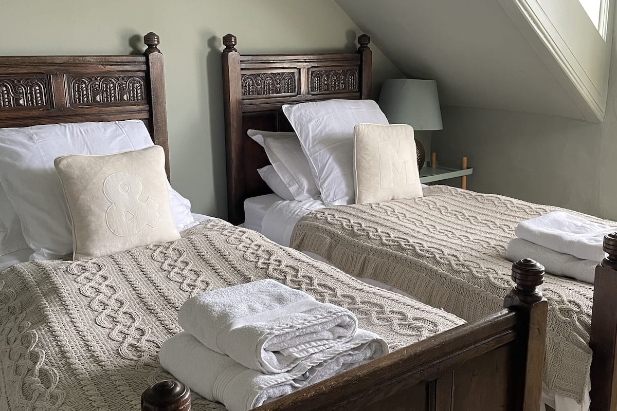 Quarto com duas camas de madeira, roupa de cama branca e uma mesa de cabeceira com abajur.