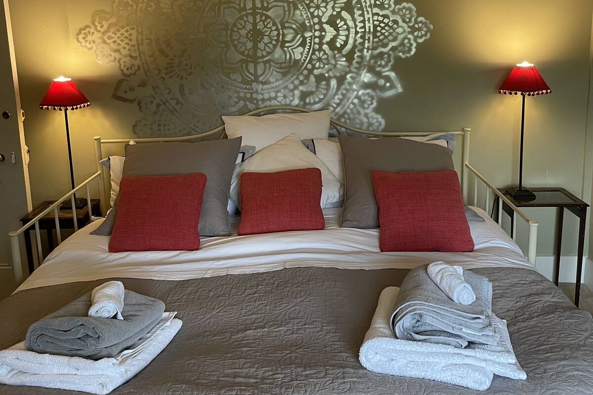 Cama bem arrumada com almofadas vermelhas e cinzentas, parede estampada ao fundo e duas luminárias de cabeceira.