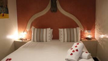 Cama arrumada com pétalas de rosa e uma toalha enrolada, dois travesseiros e luminária fixada na parede.
