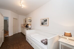 Quarto moderno com cama branca, mesa de cabeceira com abajur, uma cadeira pequena, cozinha com armários e micro-ondas, e um quadro na parede.