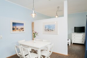 Uma sala de jantar com uma mesa e cadeiras brancas, dois quadros nas paredes, um vaso com flores e uma televisão em um aparador branco.