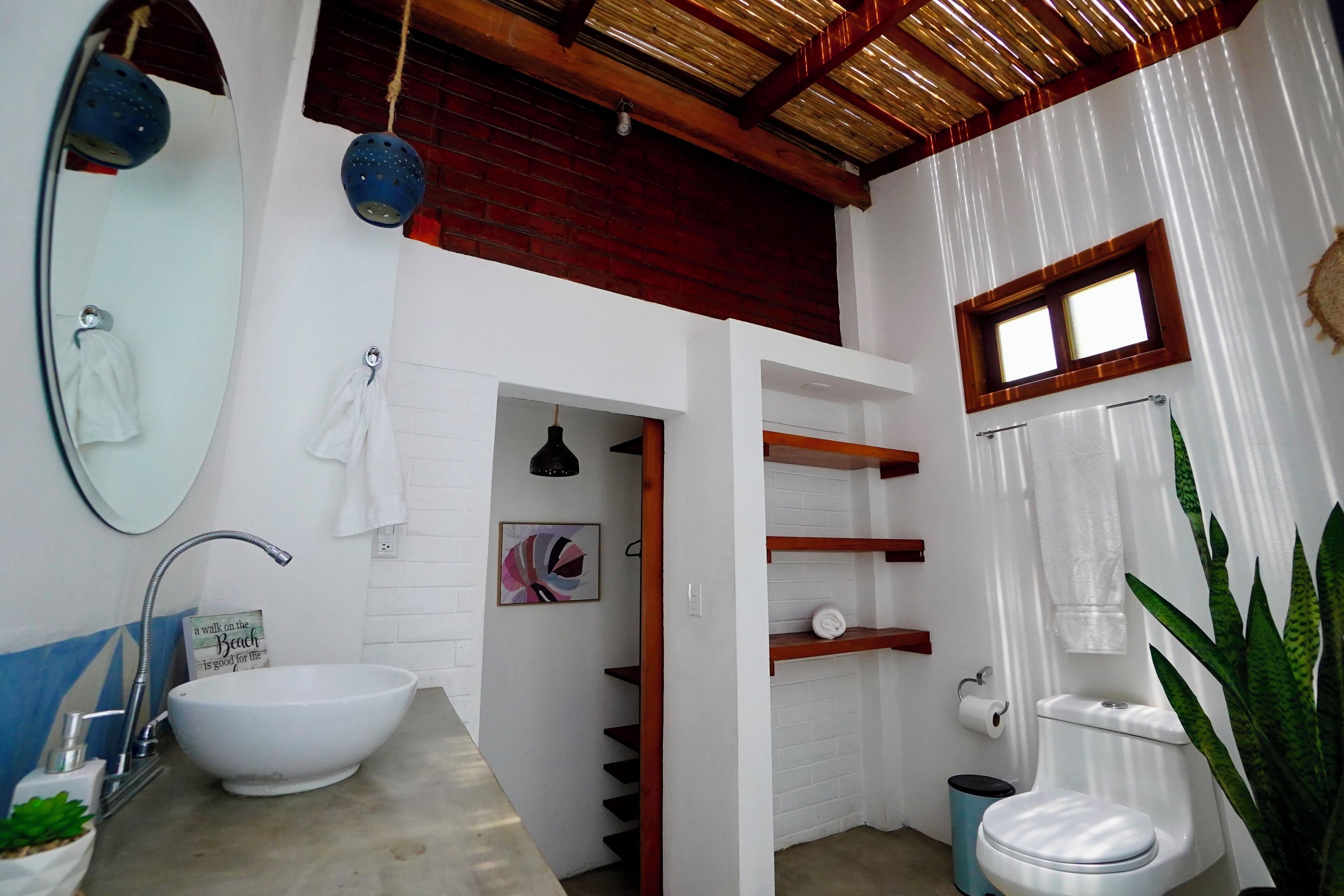 Um banheiro com pia branca, espelho, chuveiro e vaso sanitário. Há uma janela com cortinas e uma planta no canto.