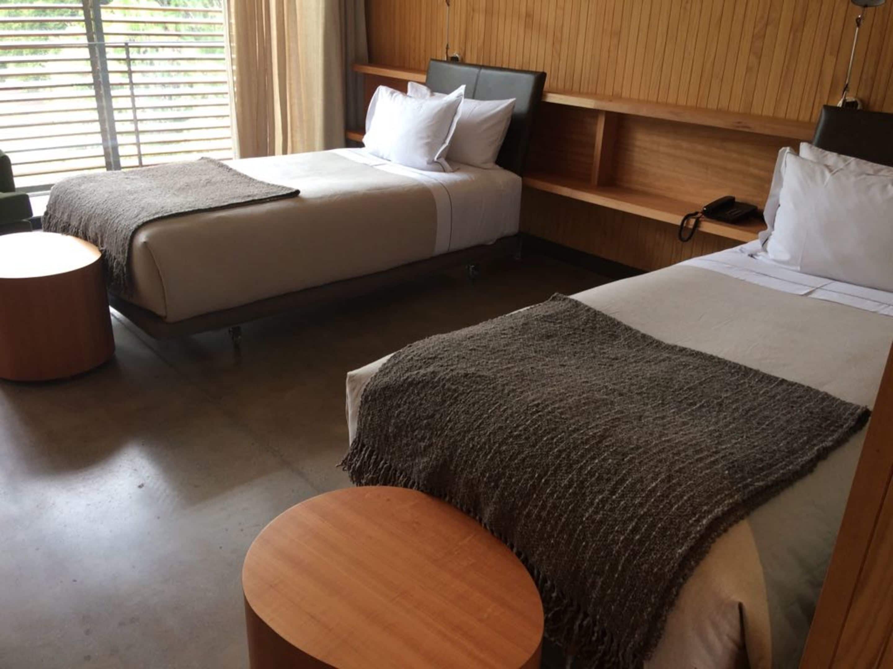 Quarto de hotel com duas camas, cabeceira de madeira, mesa de cabeceira e um banquinho redondo de madeira.