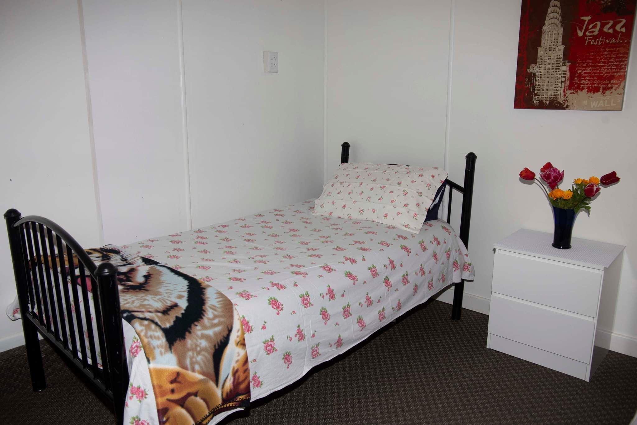 Uma cama de solteiro com colcha florida, uma mesa de cabeceira com um vaso de flores e um cartaz na parede.