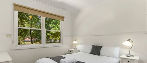 Uma cama bem arrumada com um edredom cinza e branco, duas luminárias de cabeceira, uma janela com vista para o verde e uma mesinha de cabeceira branca.
