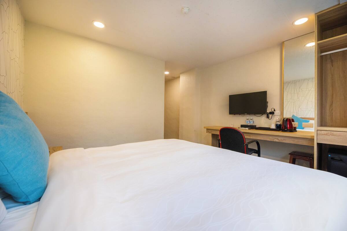 Quarto de hotel com cama, escrivaninha, cadeira, televisão e espelho.