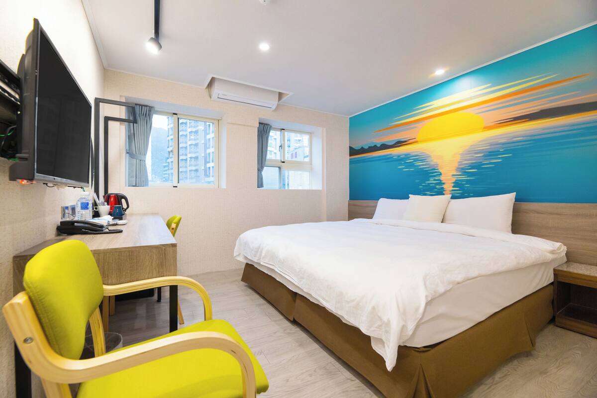 Quarto de hotel com cama, escrivaninha com cadeira, televisão e pintura de um pôr do sol.