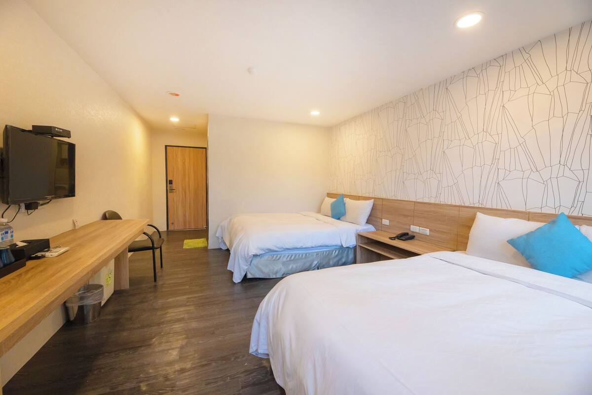 Quarto de hotel com duas camas, uma escrivaninha, uma televisão e cabeceira de madeira.
