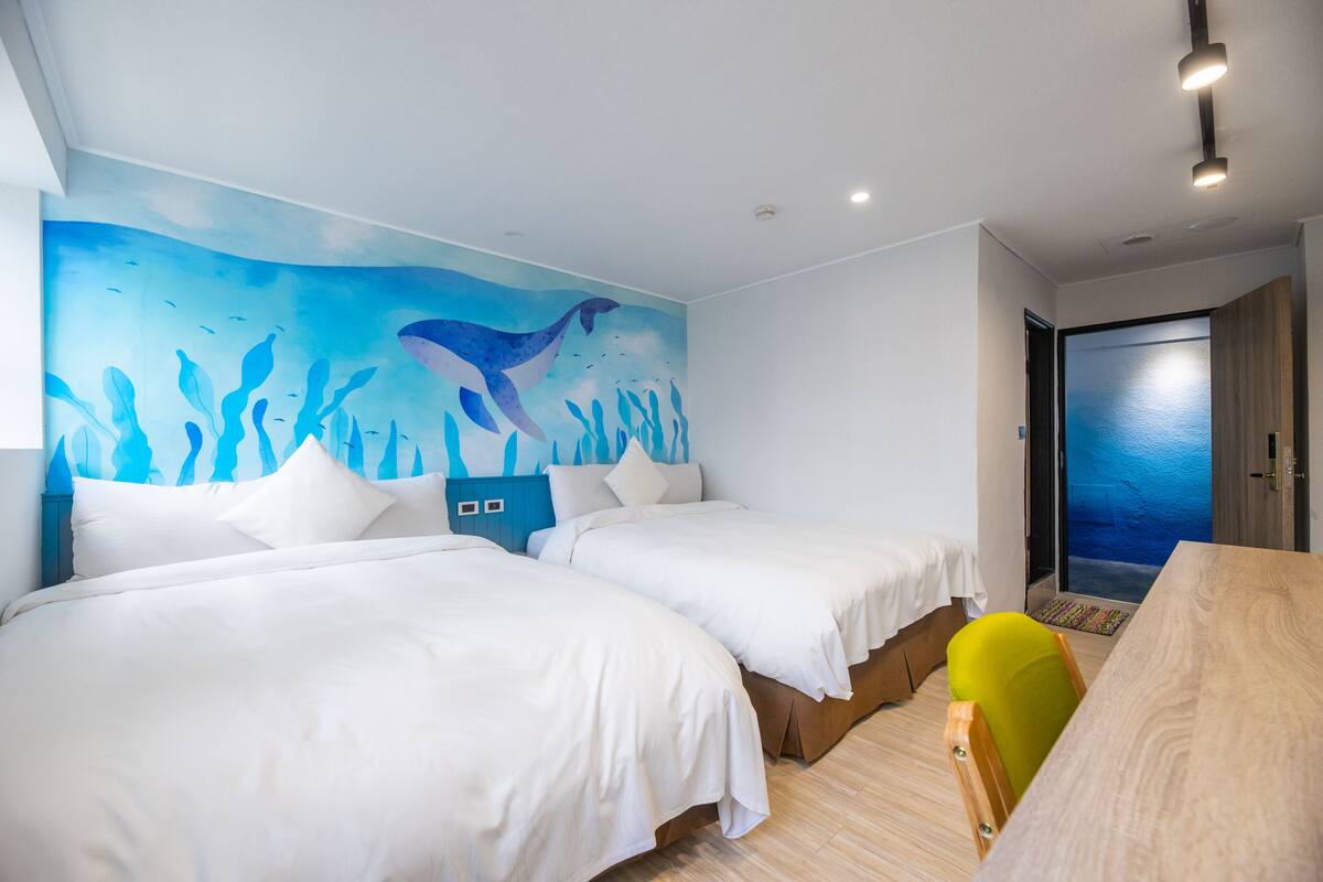 Quarto de hotel com duas camas, uma mesa de jantar de madeira e um mural na parede retratando uma cena subaquática.