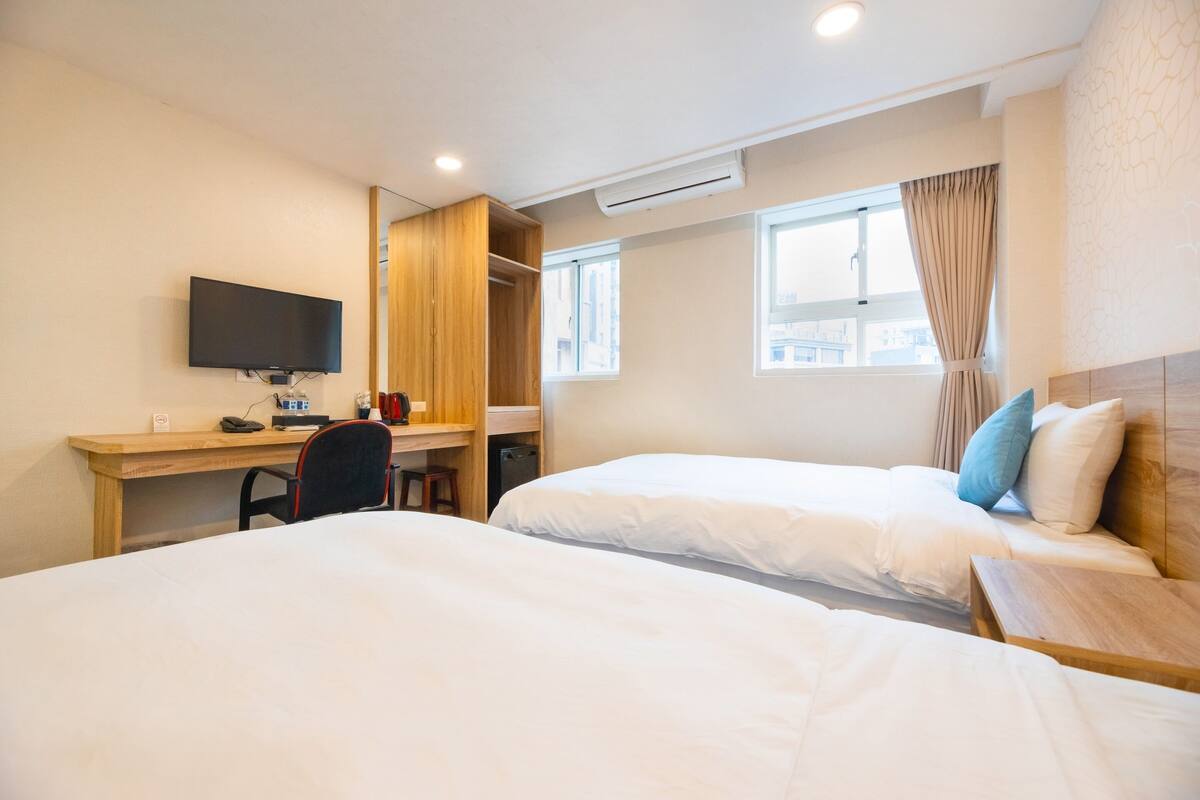Quarto de hotel com duas camas, uma escrivaninha, uma televisão e uma janela com cortinas.