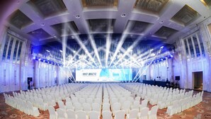 Um salão amplo com fileiras de cadeiras brancas, um palco com uma grande tela e um teto com detalhes elaborados.