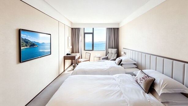 Quarto de hotel com duas camas, uma escrivaninha, uma cadeira, televisão e janela com vista para o mar.