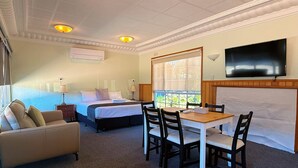 Quarto de hotel com cama, sofá, mesa de jantar com cadeiras, televisão na parede e janela com vista para vegetação.