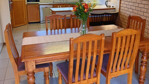 Uma área de refeições com mesa e cadeiras de madeira, um vaso com flores e uma cozinha ao fundo.