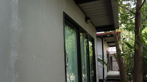 Um corredor externo coberto com portas de vidro que dão para uma varanda, ladeado por árvores.