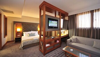 Quarto de hotel com uma cama, sofá, televisão e escrivaninha com luminária.
