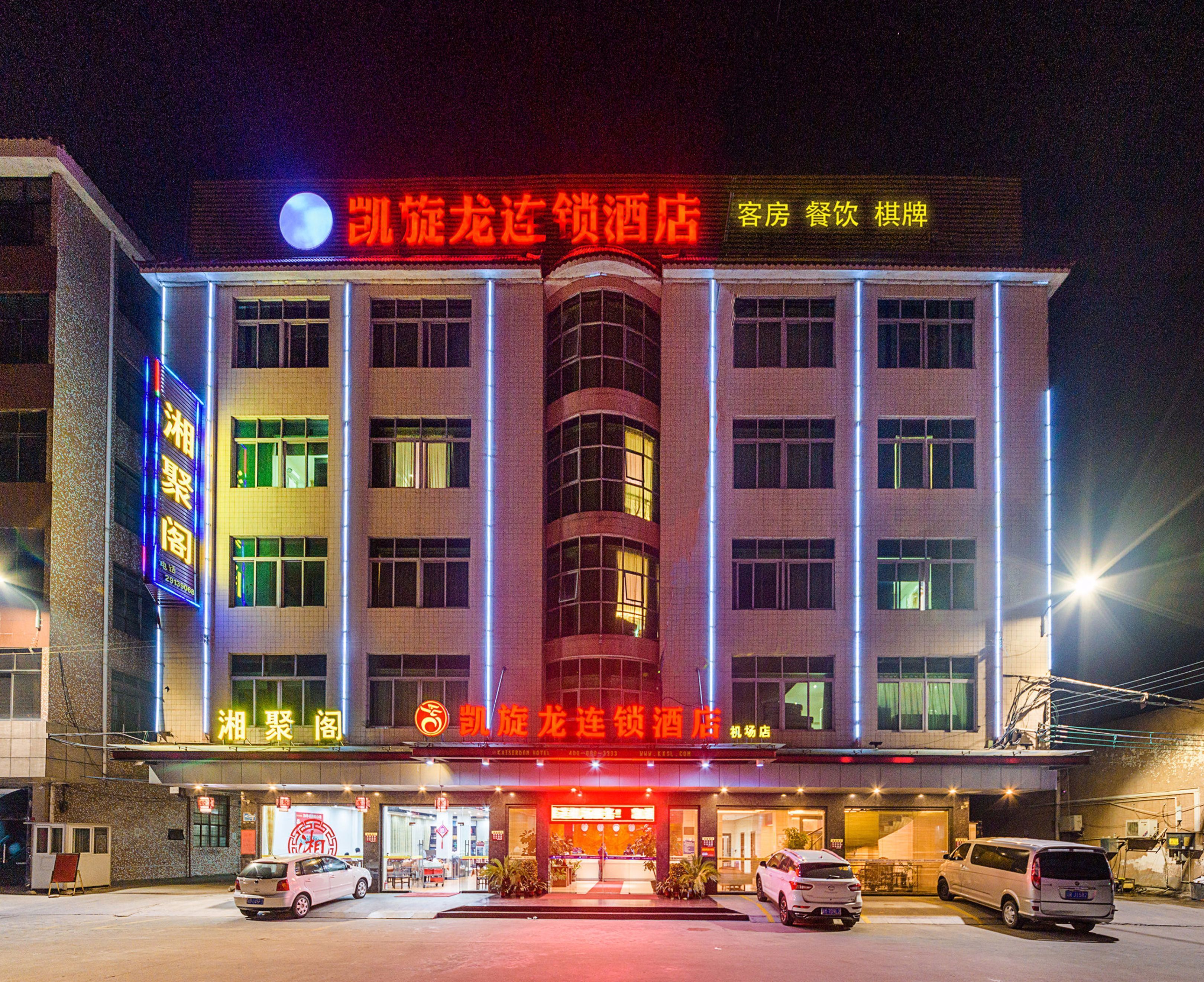 Um hotel com vários andares, entrada central e estacionamento com diversos carros.