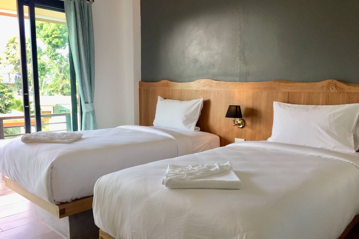Quarto de hotel com duas camas, cabeceiras de madeira, roupa de cama branca, luminária fixada na parede e vista para o verde lá fora.