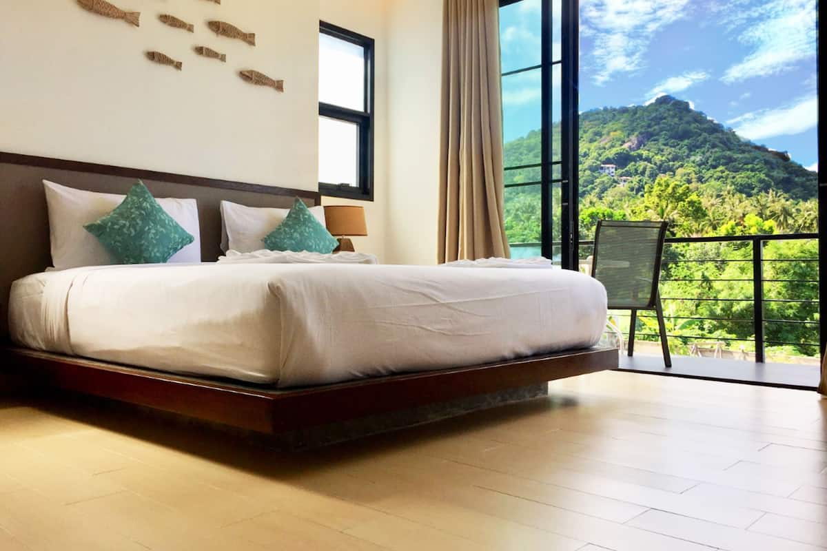 Uma cama grande com cabeceira de madeira, várias almofadas e vista para uma paisagem verde exuberante através de uma janela ampla.