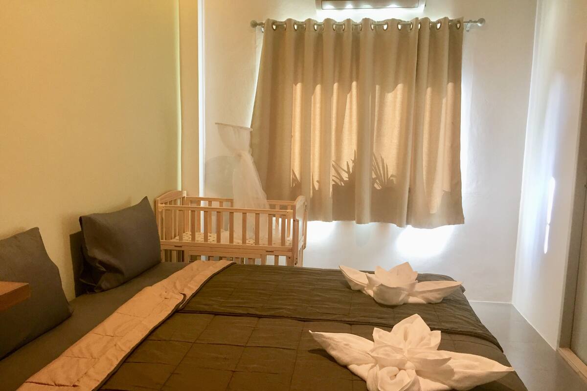 Quarto de hotel com uma cama, berço de madeira e janela com cortinas.
