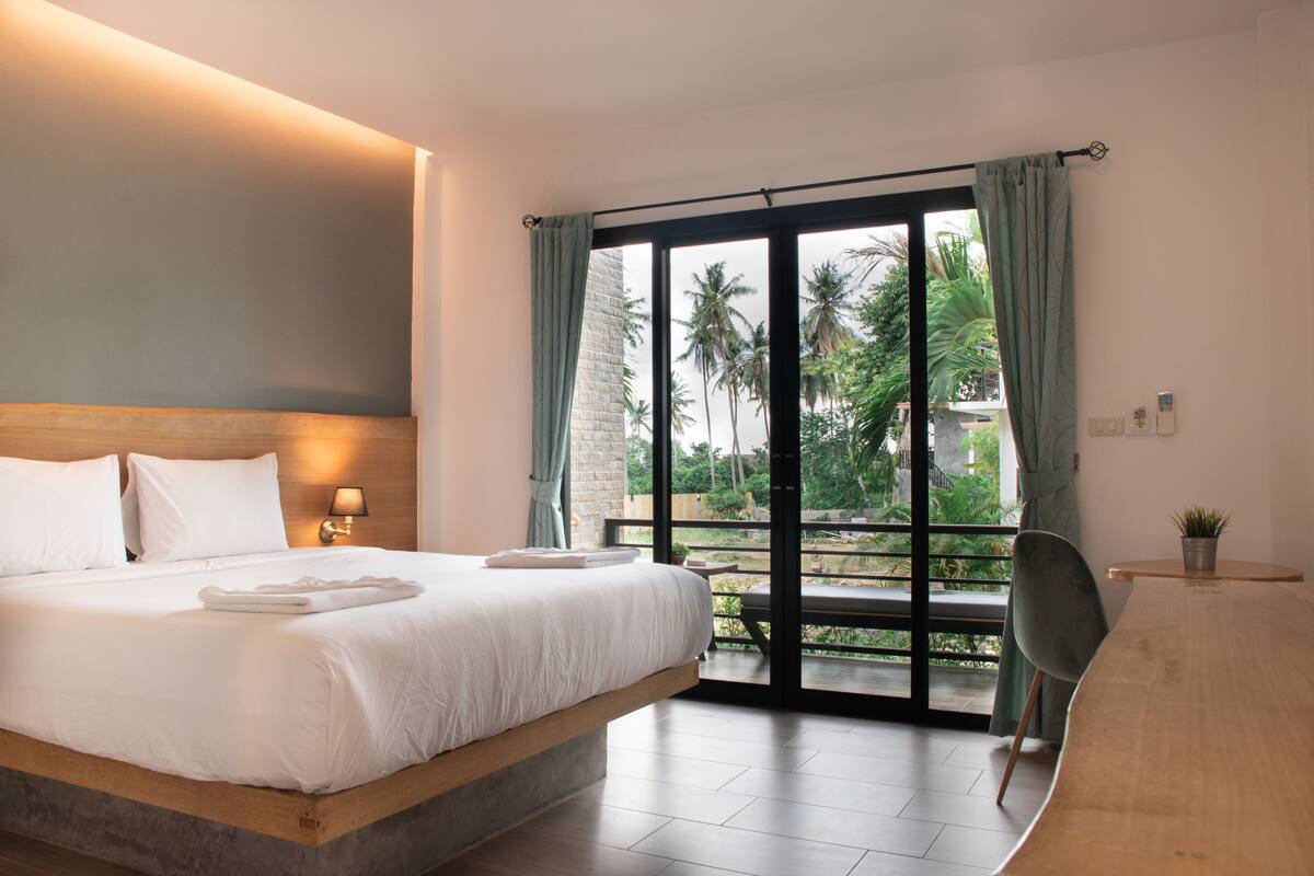 Quarto de hotel com uma cama grande, uma escrivaninha e vista para palmeiras pela janela.
