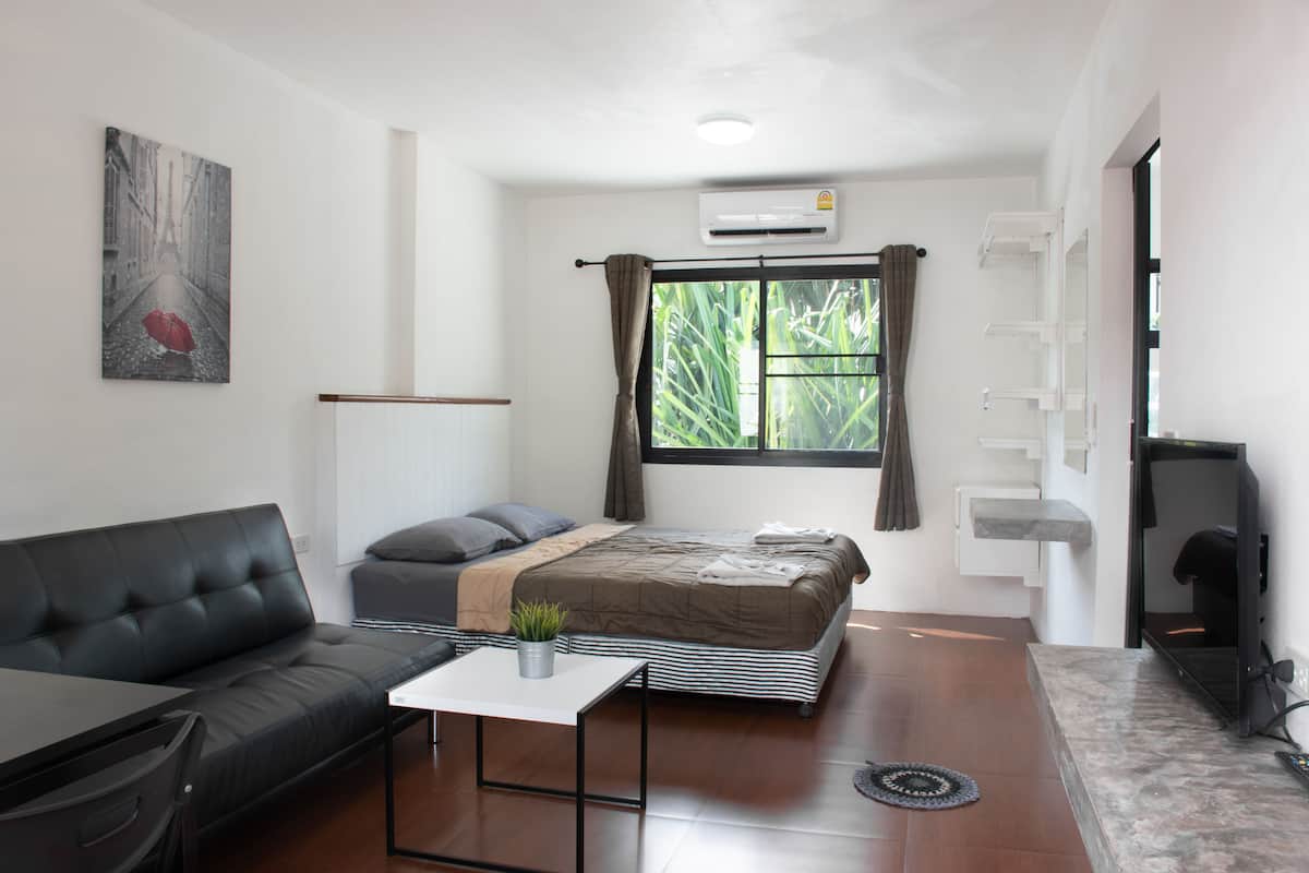 Quarto compacto com cama, sofá, uma mesinha com uma planta, televisão e janela com cortinas.