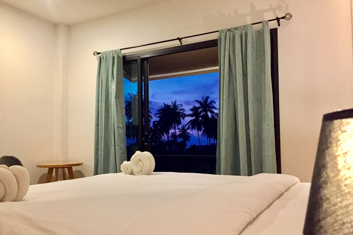 Quarto de hotel com cama, uma mesinha e janela com cortinas.