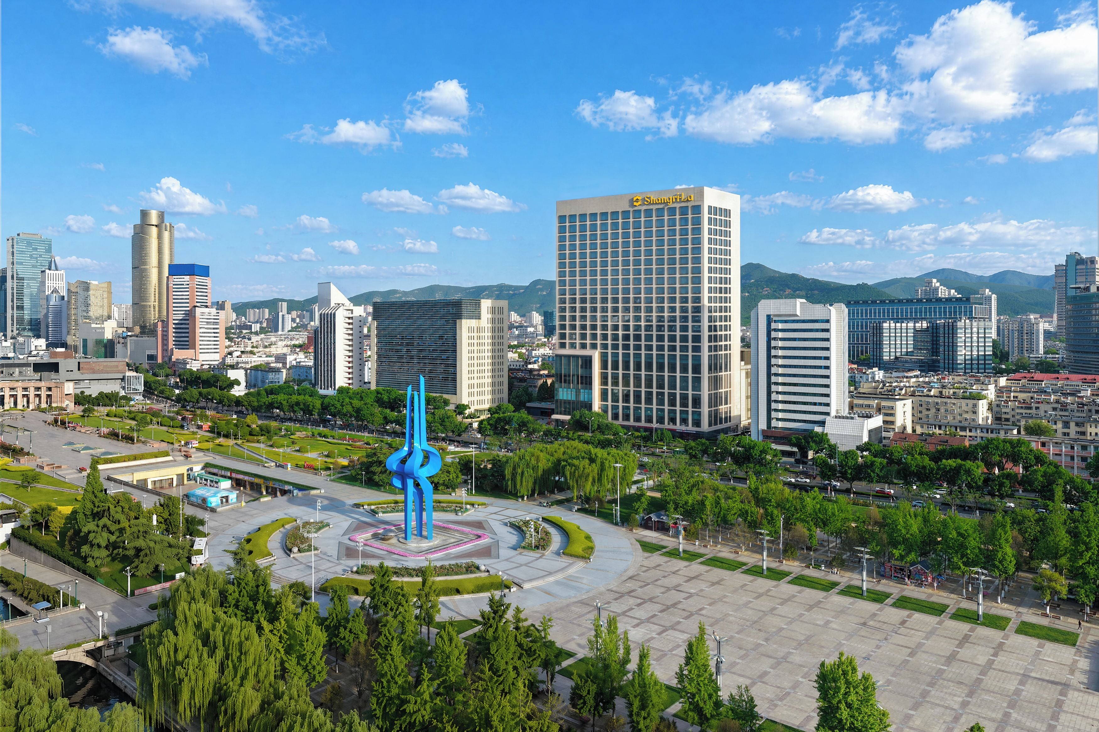 Paisagem urbana com edifícios modernos, uma escultura azul no centro e um parque com uma praça circular.