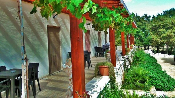 Uma área de estar coberta ao ar livre, com mesas e cadeiras, cercada por vegetação e uma pérgola de madeira.