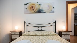 Cama bem arrumada com colcha florida, duas mesas de cabeceira com abajures e um quadro com rosas na parede.