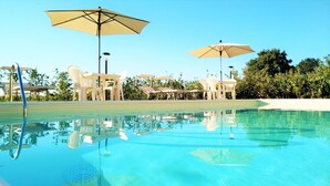 Piscina cristalina com espreguiçadeiras e guarda-sóis, cercada por vegetação e um céu azul.
