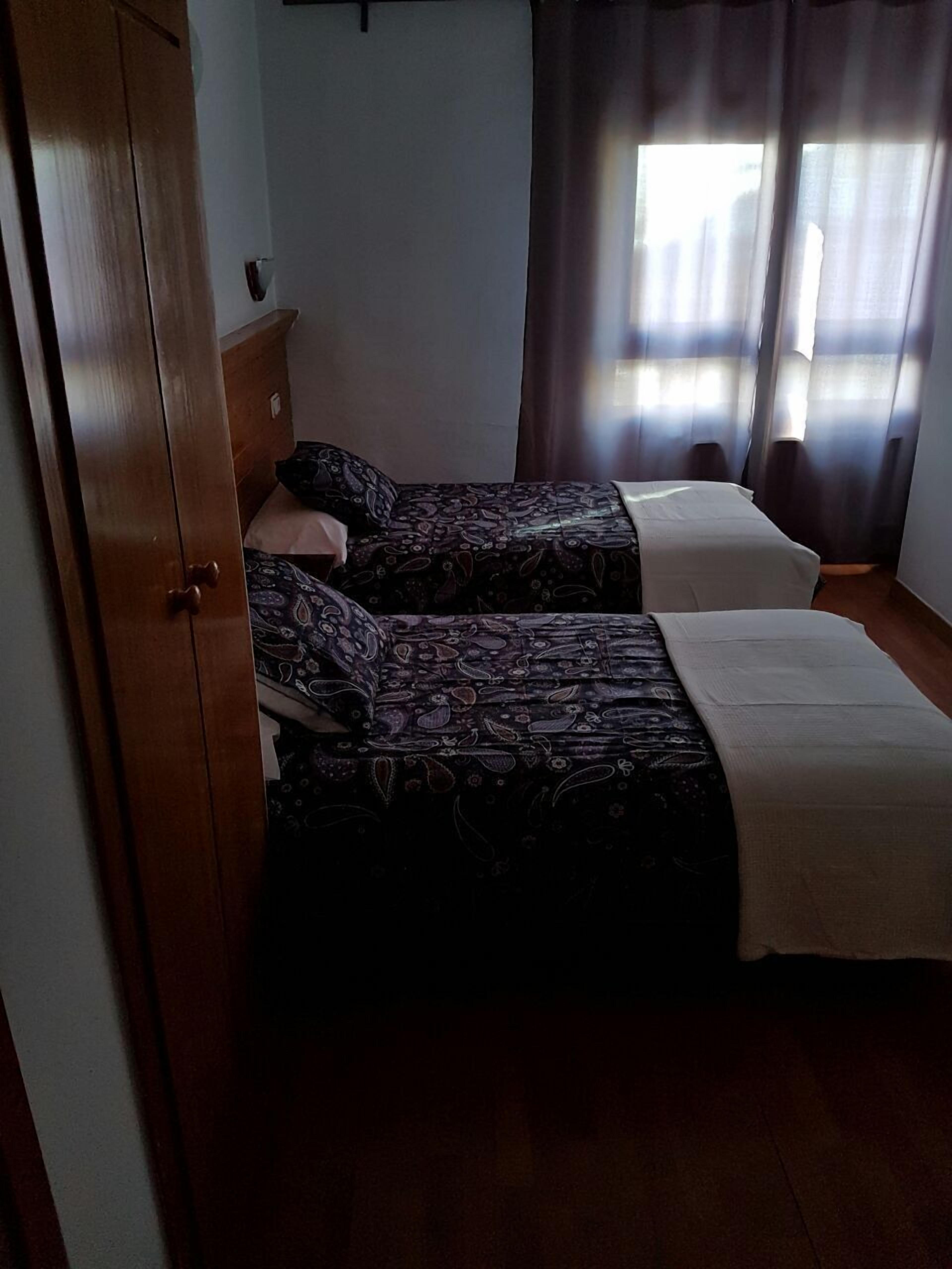 Quarto com duas camas, uma com colcha estampada e outra com colcha lisa. Um guarda-roupa de madeira à esquerda e uma janela com cortinas à direita.