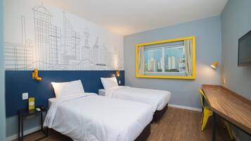 Quarto de hotel com duas camas, um mural com vista para a cidade, janela com moldura amarela e uma escrivaninha com cadeira.